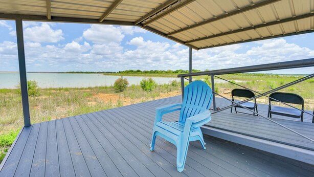 Um deck coberto com vista para um lago, uma cadeira de plástico azul e duas cadeiras pretas.