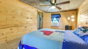 Quarto de cabana de madeira com cama, ventilador de teto e janela com cortinas.
