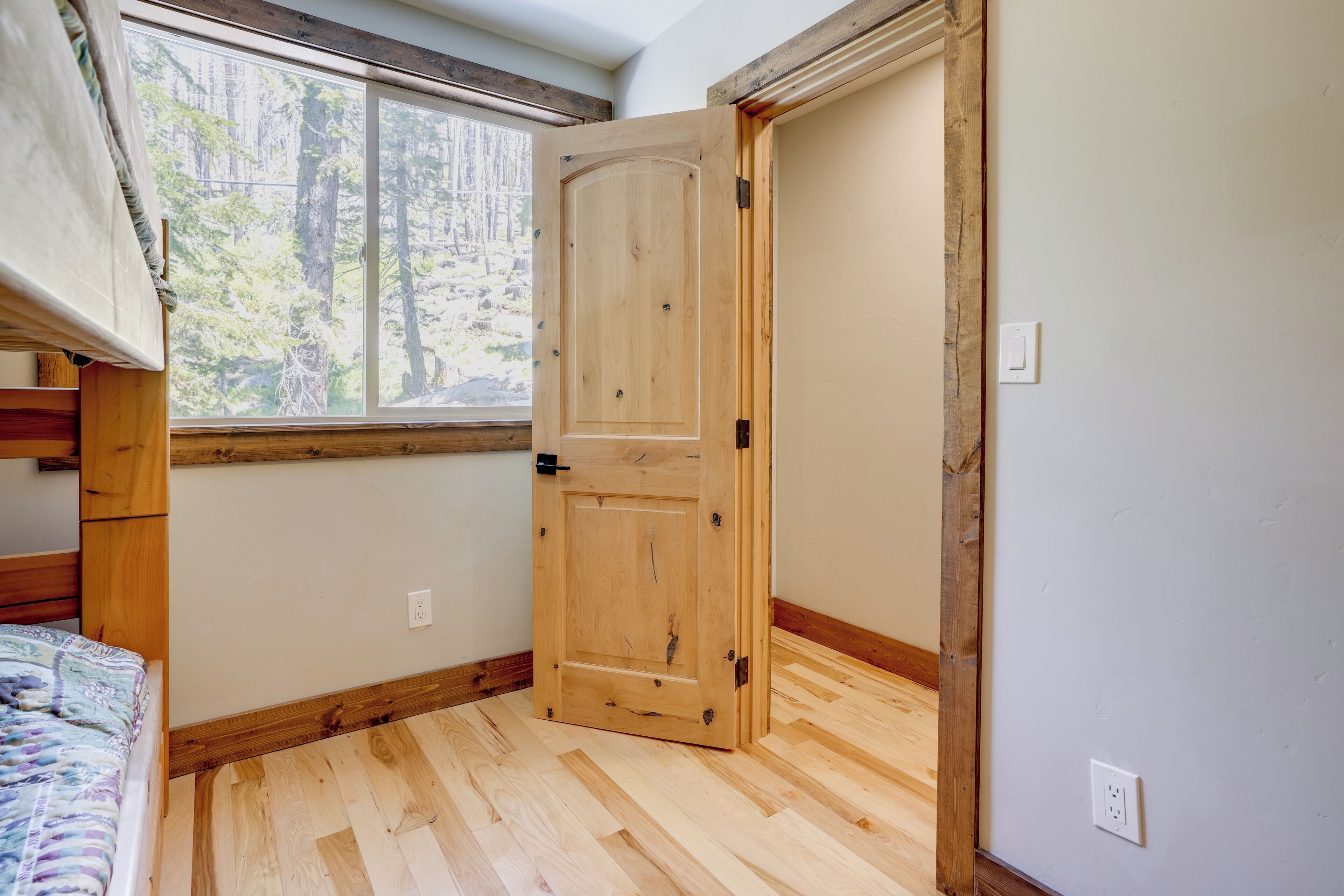 Quarto com porta de madeira, janela com vista para uma floresta, cama com colcha estampada e piso de madeira.