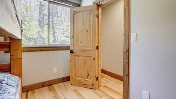 Quarto com porta de madeira, janela com vista para uma floresta, cama com colcha estampada e piso de madeira.