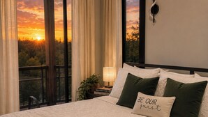 Quarto com cama grande, mesa de cabeceira com abajur e vista para o pôr do sol pela janela.