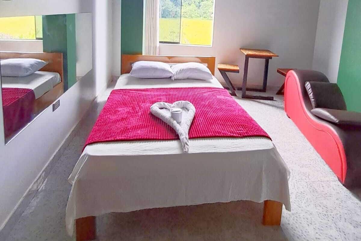 Quarto de hotel com cama, cadeira, mesa pequena e janela com vista para o verde.