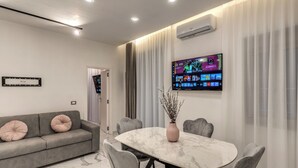 Uma sala de estar moderna com um sofá cinzento, uma mesa de jantar de mármore, cadeiras cinzas, uma TV montada na parede e um aparelho de ar condicionado.