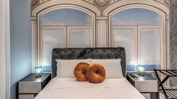 Quarto com cabeceira estofada, duas luminárias de apoio e vista para o mar através de janelas em arco.