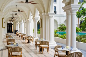 Um espaço de jantar de hotel com colunas brancas, mesas redondas, cadeiras de vime e ventoinhas de teto.