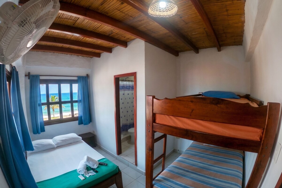 Quarto com cama de solteiro, uma cama beliche, janela com vista para a praia, ventilador de teto e porta que dá para outro quarto.