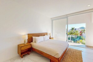 Quarto com uma cama grande, mesa de cabeceira, luminária e vista para palmeiras e edifícios.