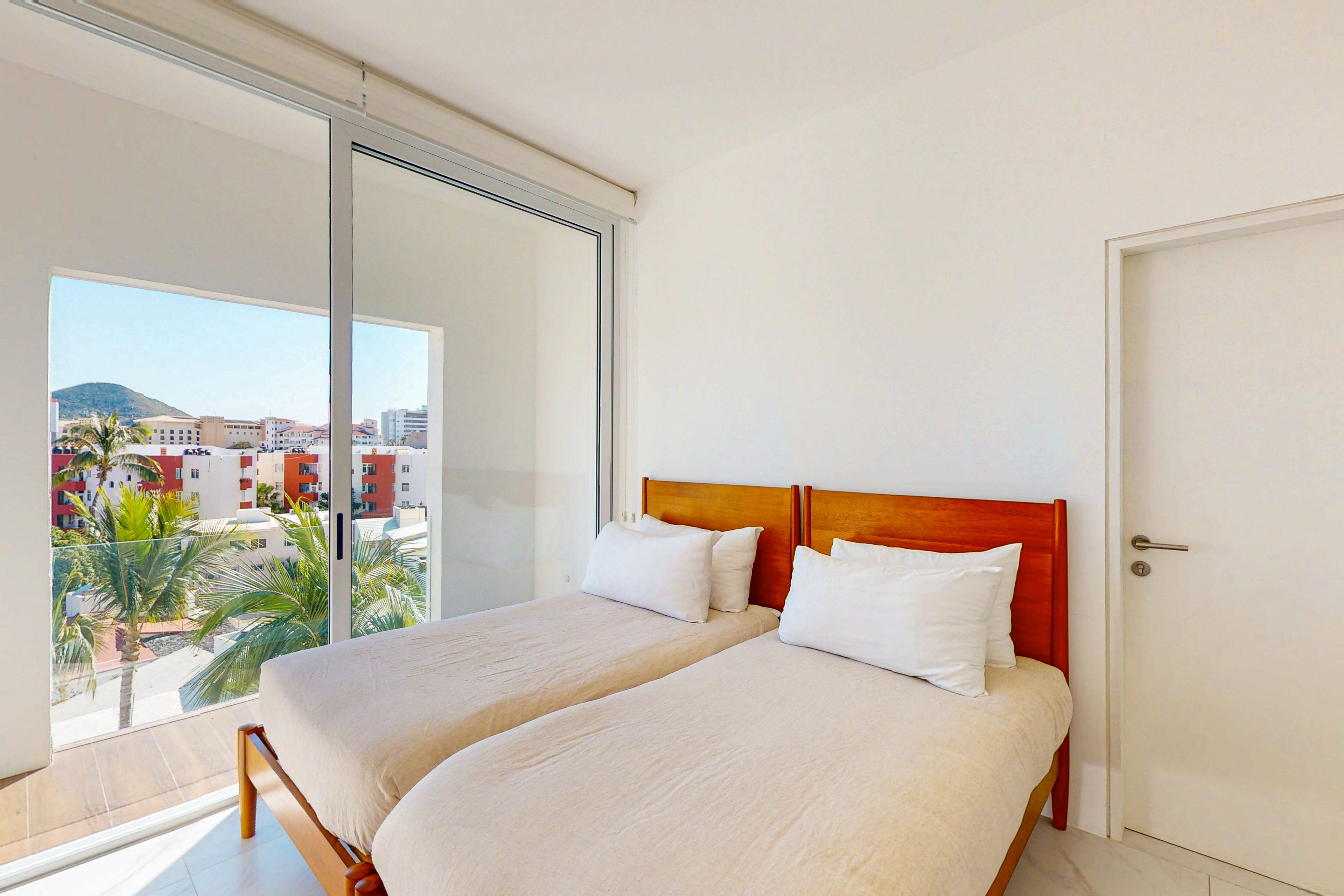 Quarto com duas camas, janelas amplas e vista para edifícios e palmeiras.