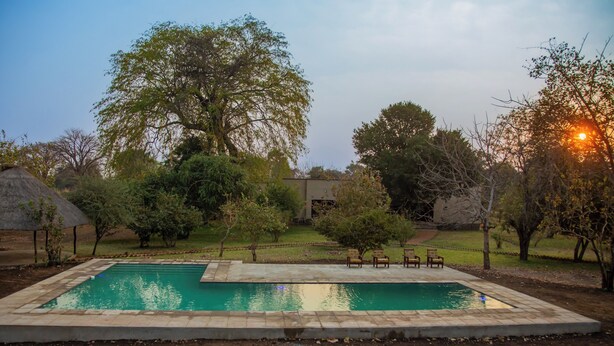 Uma piscina retangular com borda de pedra, rodeada por árvores e uma estrutura com telhado de palha.
