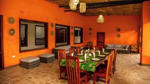 Uma área de jantar com mesa posta para seis pessoas, rodeada por cadeiras de madeira, e um sofá ao fundo.