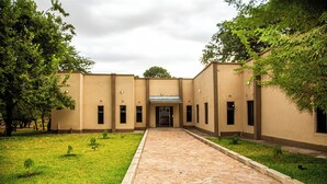 Um edifício com entrada central, um caminho pavimentado e uma área ajardinada com plantas pequenas.
