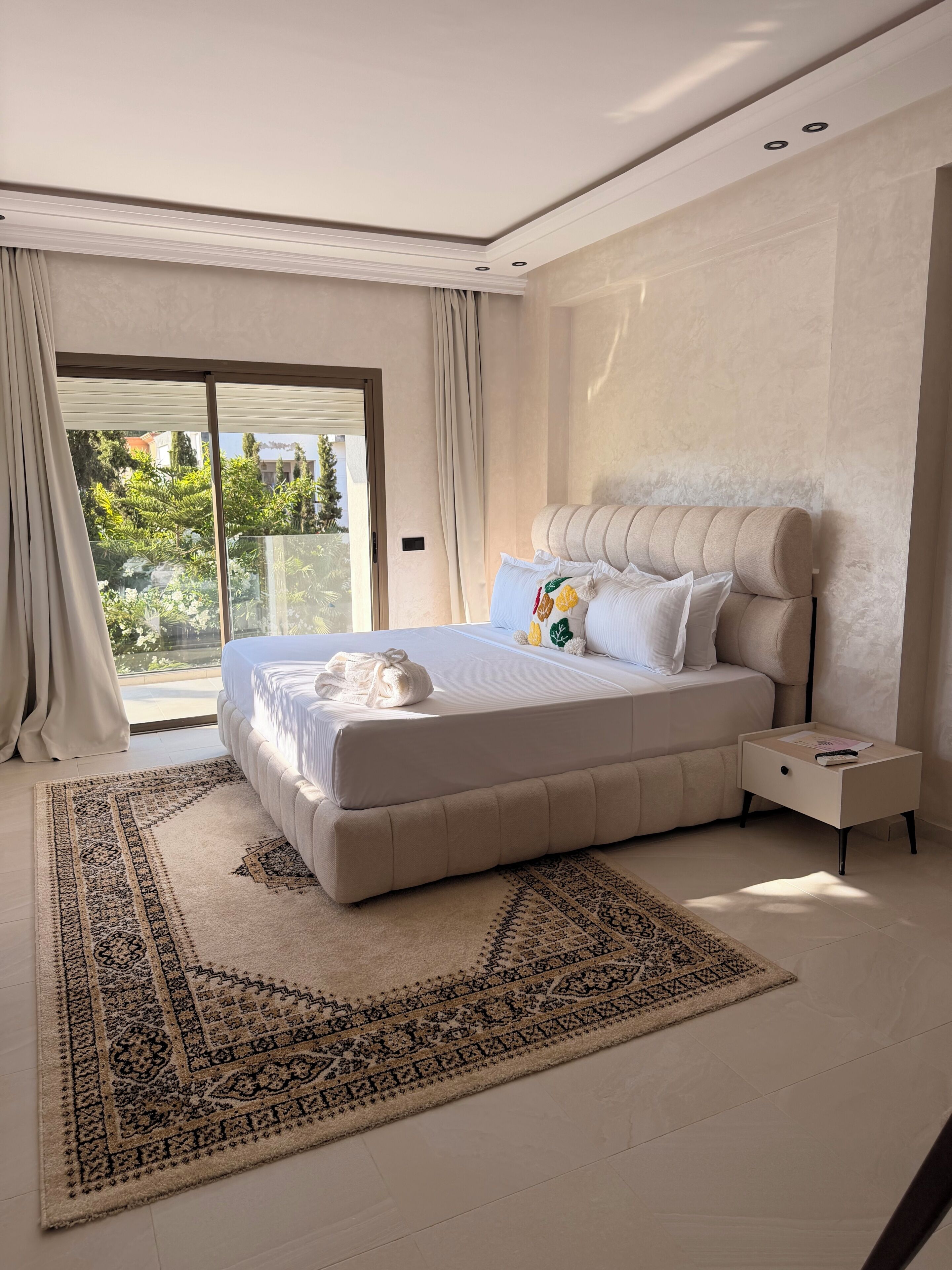 Um quarto moderno com uma cama grande, uma mesa de cabeceira e vista para o verde pela janela.