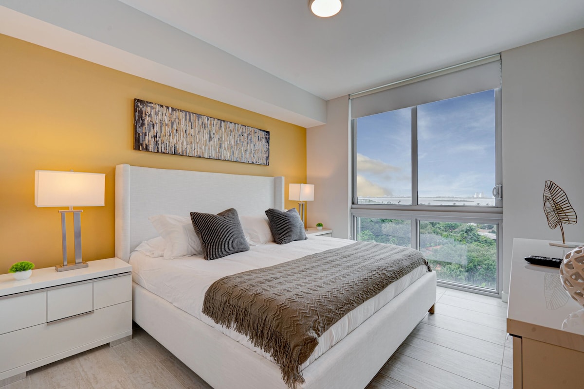 Quarto moderno com uma cama grande, duas mesas de cabeceira, um criado-mudo, uma janela com vista e uma luminária de teto.