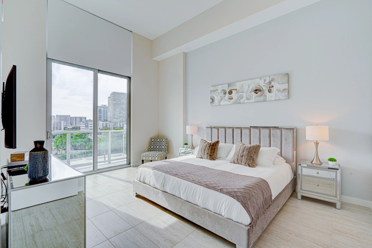 Quarto moderno com cama grande, vista para a cidade, televisão e um vaso decorativo.