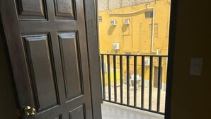 Uma porta de madeira escura fechada dá para uma varanda, com um edifício amarelo ao fundo.