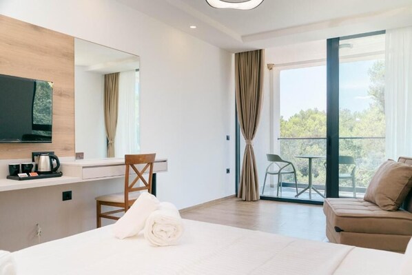 Quarto de hotel com cama, mesa de trabalho com cafeteira, poltrona, sofá, varanda com vista e televisão na parede.