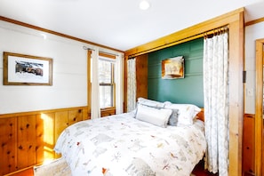 Um quarto com cama, paredes de madeira, uma parede verde de destaque, um quadro e uma janela com cortinas.