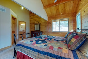 Quarto com teto de madeira, uma janela grande, cama com colcha, cómoda e espelho.