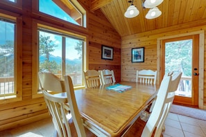 Uma cabana de madeira com mesa de jantar, cadeiras e grandes janelas com vista para as montanhas.