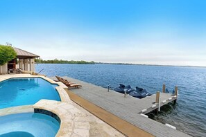 Piscina com deck de madeira e jet skis, área coberta com cadeiras e vista para a água.