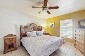 Quarto com cama, mesa de cabeceira de madeira, cômoda, ventilador de teto e janela com persianas.
