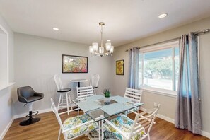Uma sala de jantar com mesa de vidro, cadeiras brancas, uma cadeira preta, um candelabro e uma janela com cortinas.