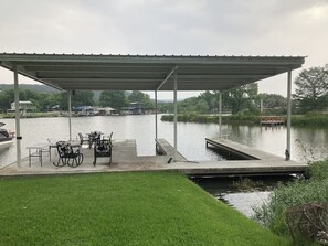Um cais coberto com cadeiras e uma mesa, uma área gramada e um lago calmo com barcos ao fundo.