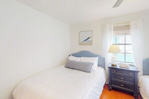 Um quarto com cama, mesa de cabeceira com abajur, janela com cortinas e um quadro na parede.
