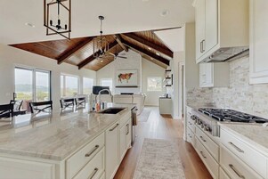 Uma cozinha espaçosa com uma ilha grande, um espaço de refeições com mesa e cadeiras, e vista para o exterior através de janelas amplas.