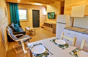 Uma cabana de madeira com sofá, mesa de jantar para dois, televisão e cozinha.