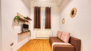 Um espaço de estar compacto com um sofá marrom, piso de madeira, radiador branco e uma janela com cortinas marrons.