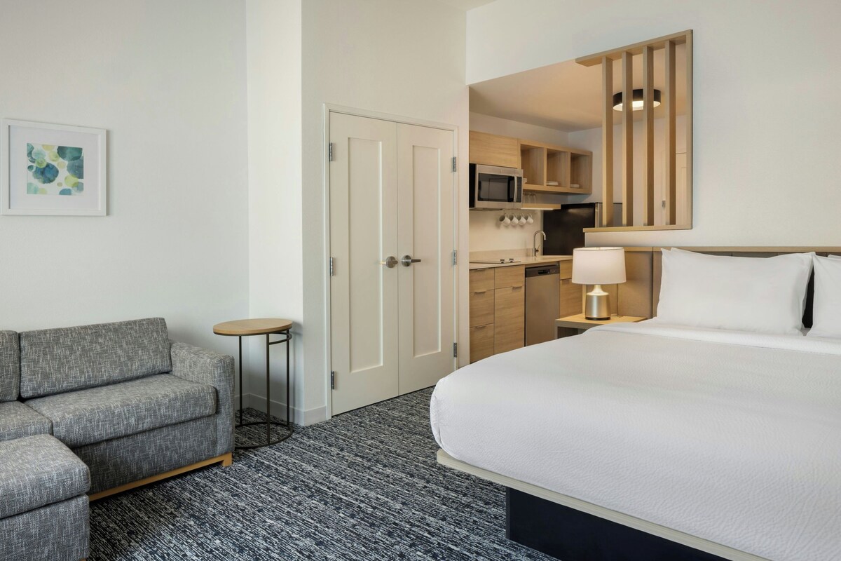 Quarto de hotel com cama, sofá, mesa pequena, micro-ondas, frigorífico e cozinha equipada com armários e luminária suspensa.