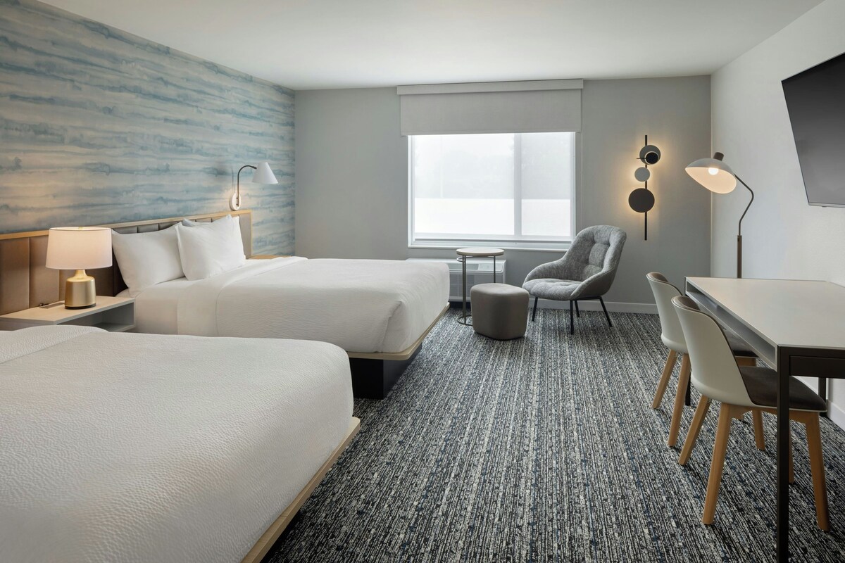 Quarto de hotel com duas camas, uma mesa, uma cadeira, um abajur e luminária na parede.