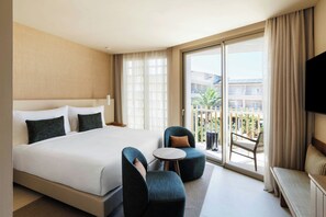 Quarto de hotel com uma cama grande, duas poltronas, uma pequena mesa e uma varanda com vista para edifícios.