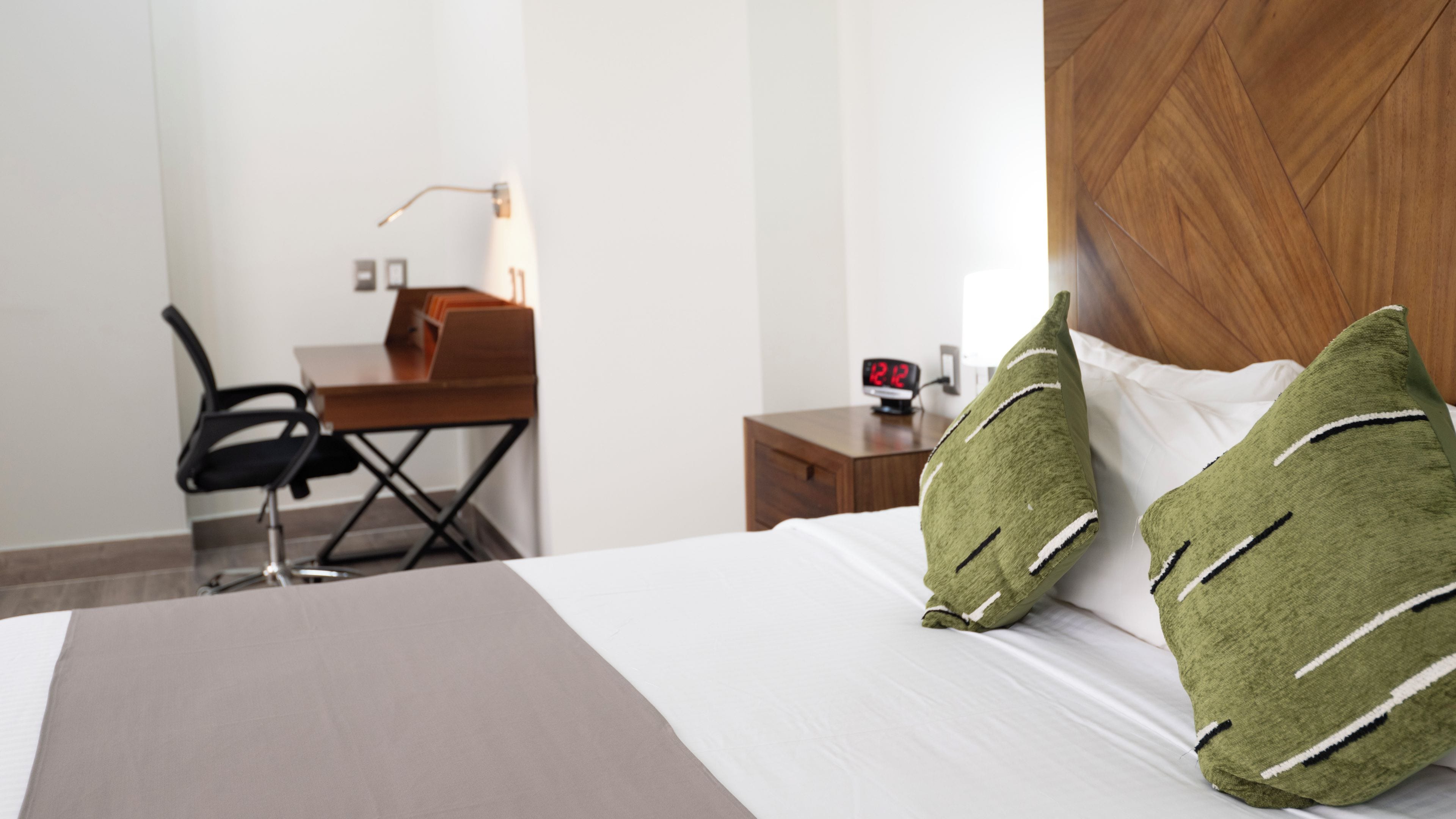Quarto de hotel com cama, escrivaninha, cadeira, mesa de cabeceira e relógio digital.
