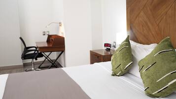 Quarto de hotel com cama, escrivaninha, cadeira, mesa de cabeceira e relógio digital.