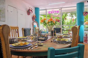 Uma área de refeições com uma mesa redonda posta para uma refeição, uma grande janela com vista para o verde, e um letreiro de neon com a inscrição "EL ÉXITO SUMA".