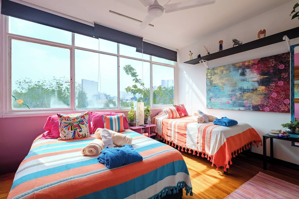 Quarto com duas camas, janelas amplas, ventoinha no teto e um quadro na parede.