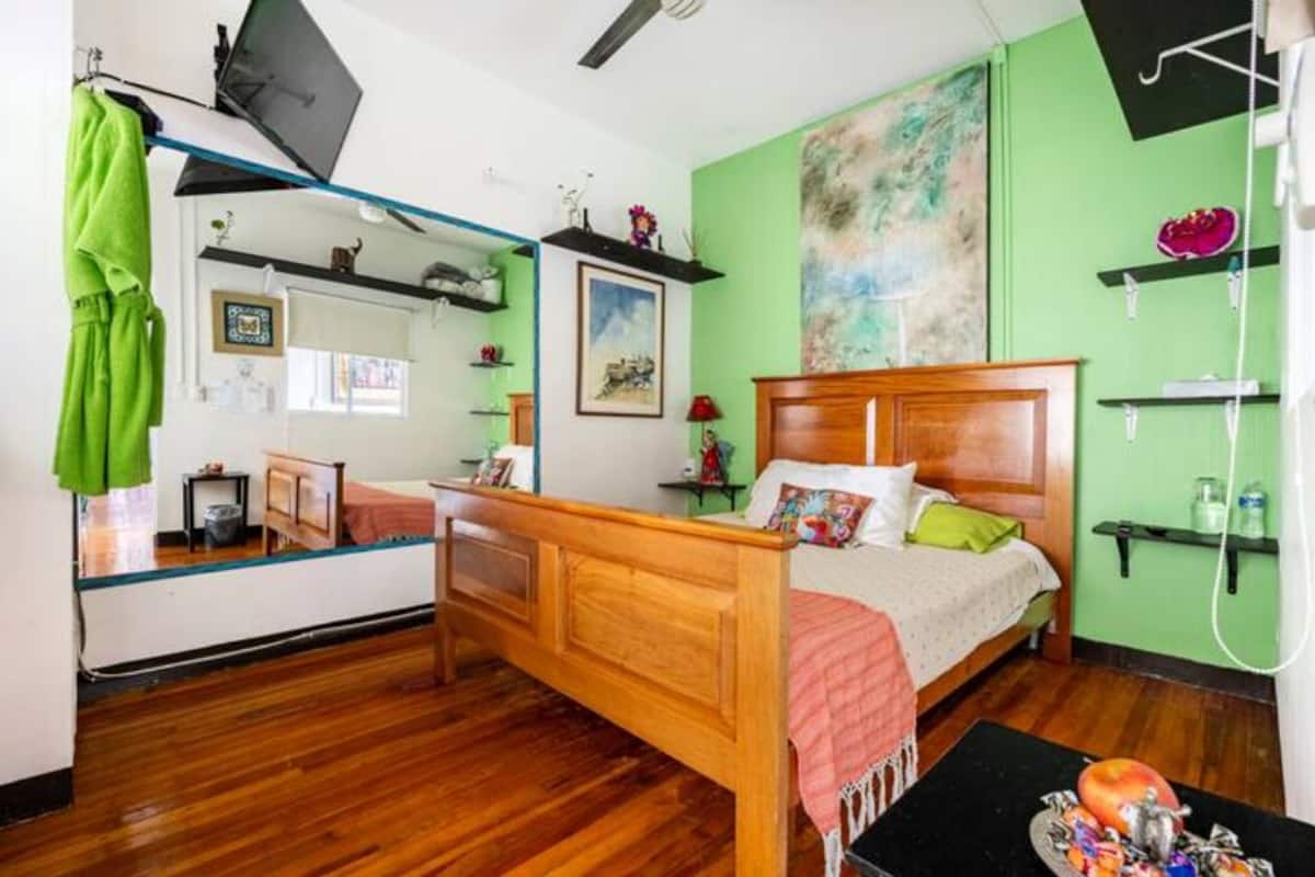 Quarto com cama de madeira, paredes verdes, um espelho e uma mesinha com abajur.