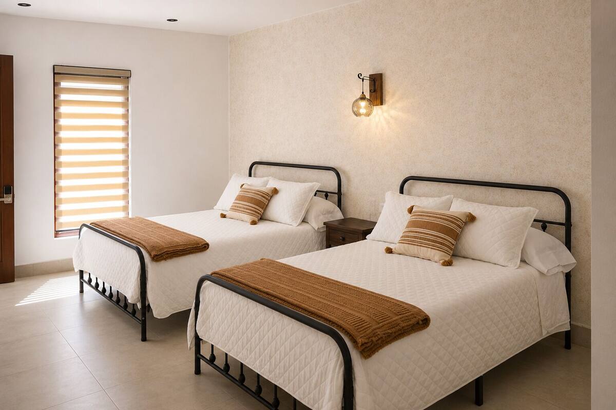 Duas camas de solteiro com cabeceiras, um candeeiro fixado na parede e uma janela com persianas.
