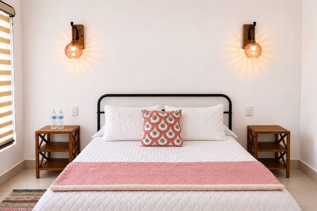 Uma cama com colcha branca e rosa, duas mesas de cabeceira de madeira, um encosto de madeira e duas luminárias fixadas na parede.