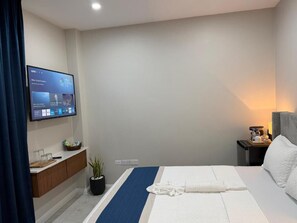 Quarto de hotel com cama, televisão fixada na parede, mesa de cabeceira com abajur e um pequeno vaso de planta.