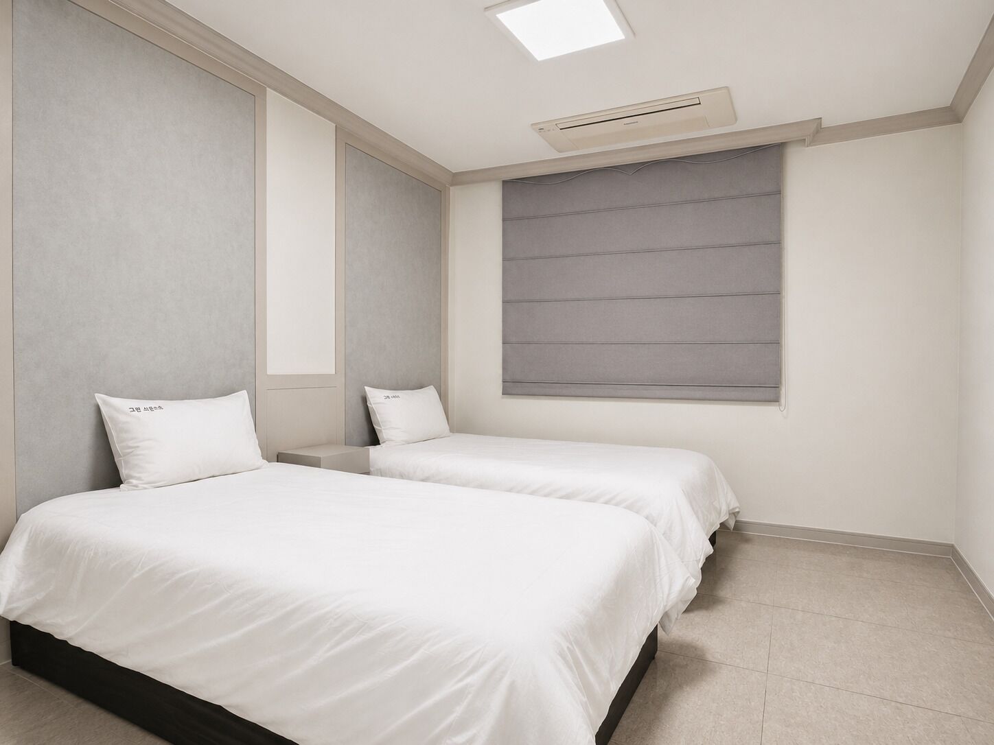 Quarto de hotel moderno com duas camas, uma janela grande com persianas, iluminação no teto e ar condicionado.