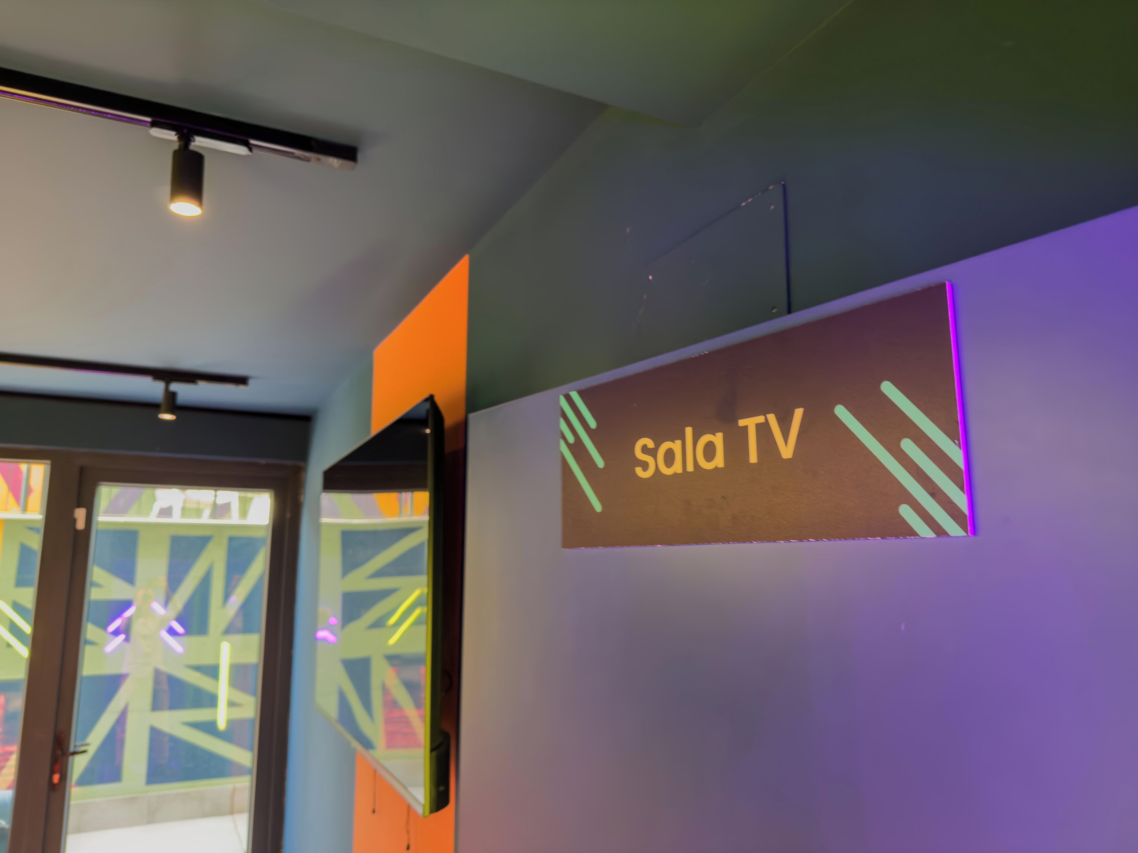 Um letreiro com a inscrição 'Sala TV' em uma parede, um lustre no teto e uma janela com padrão geométrico.
