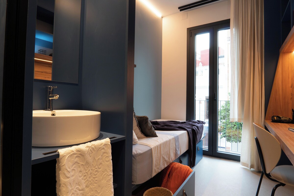Quarto de hotel moderno com uma pia grande, uma cama com almofadas, uma cadeira e vista para o exterior.