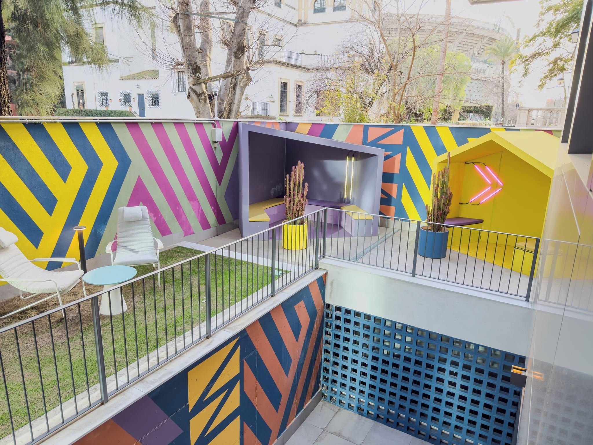 Uma área externa com vários níveis, apresentando designs geométricos coloridos na parede, assentos e plantas em vasos.