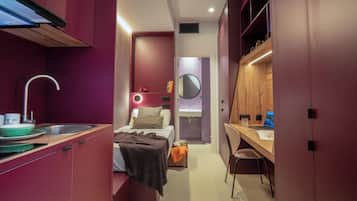 Quarto compacto com cama, mesa de cabeceira e escrivaninha com cadeira. Possui guarda-roupa embutido e uma pequena cozinha com pia e tigela.