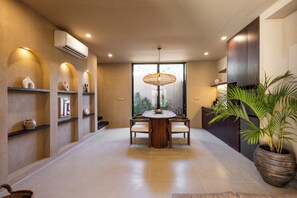 Um interior moderno com uma mesa de jantar, cadeiras, uma luminária pendente, prateleiras com objetos decorativos e uma grande planta em vaso.