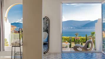 Quarto com vista para o mar, uma cadeira branca, um espelho decorativo e uma planta sobre uma mesa.