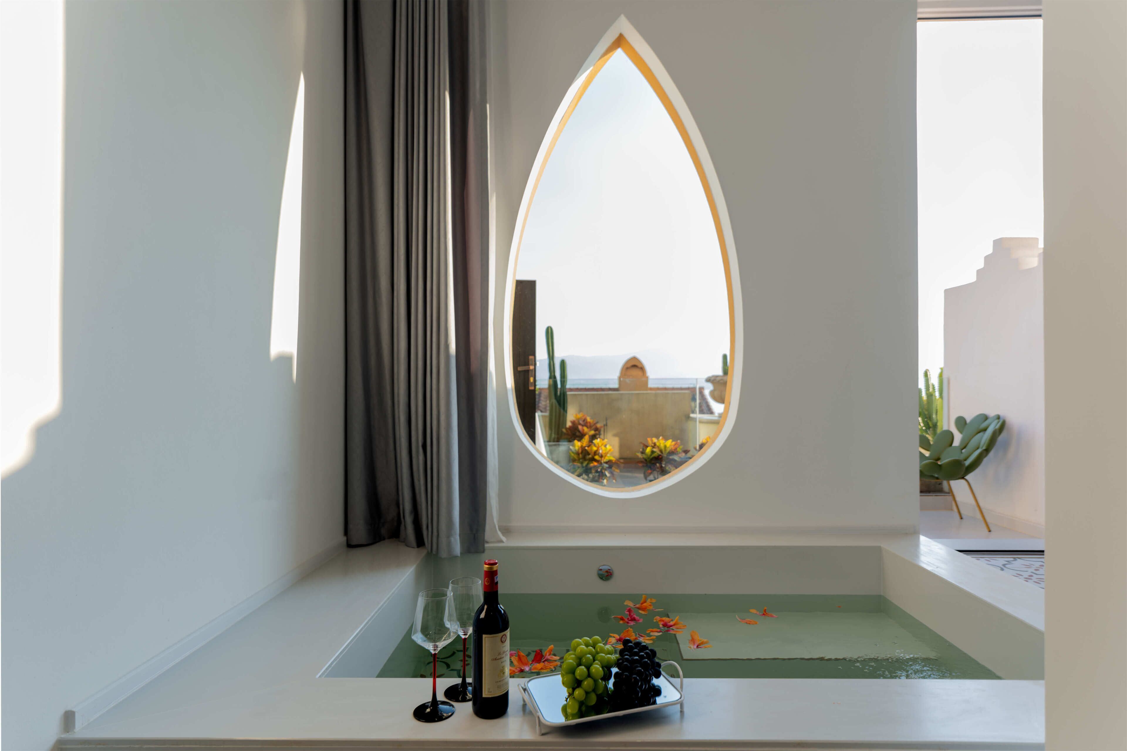 Quarto com vista pela janela, uma mesa com garrafa e copos, e uma fruteira.